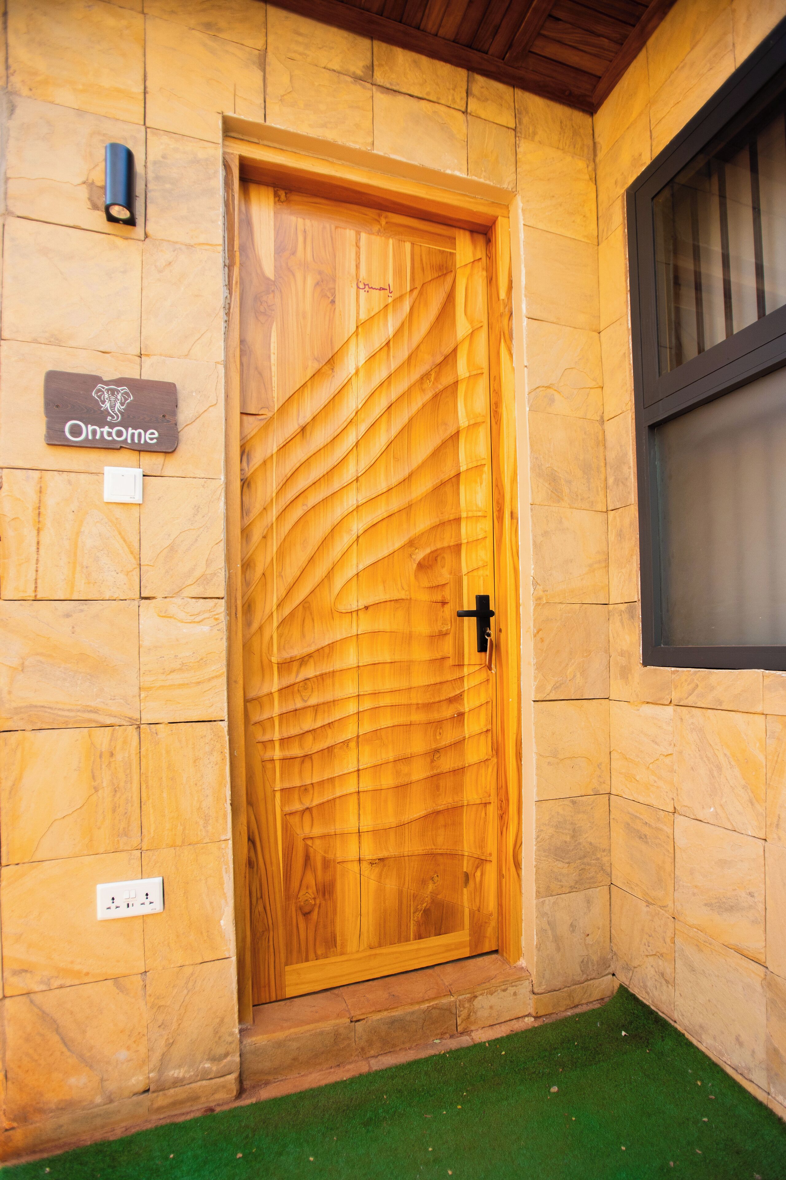 Uma porta de madeira com design único, uma placa com a palavra "Ontome" e uma janela à direita.