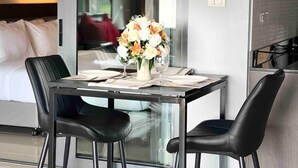 Quarto de hotel moderno com área de refeições, mesa de vidro, cadeiras pretas e um vaso de flores.