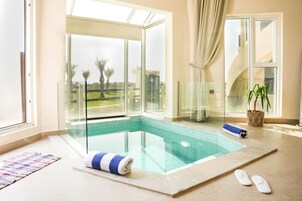 Uma piscina interior moderna com paredes de vidro, uma toalha azul e branca, e um par de chinelos no chão.