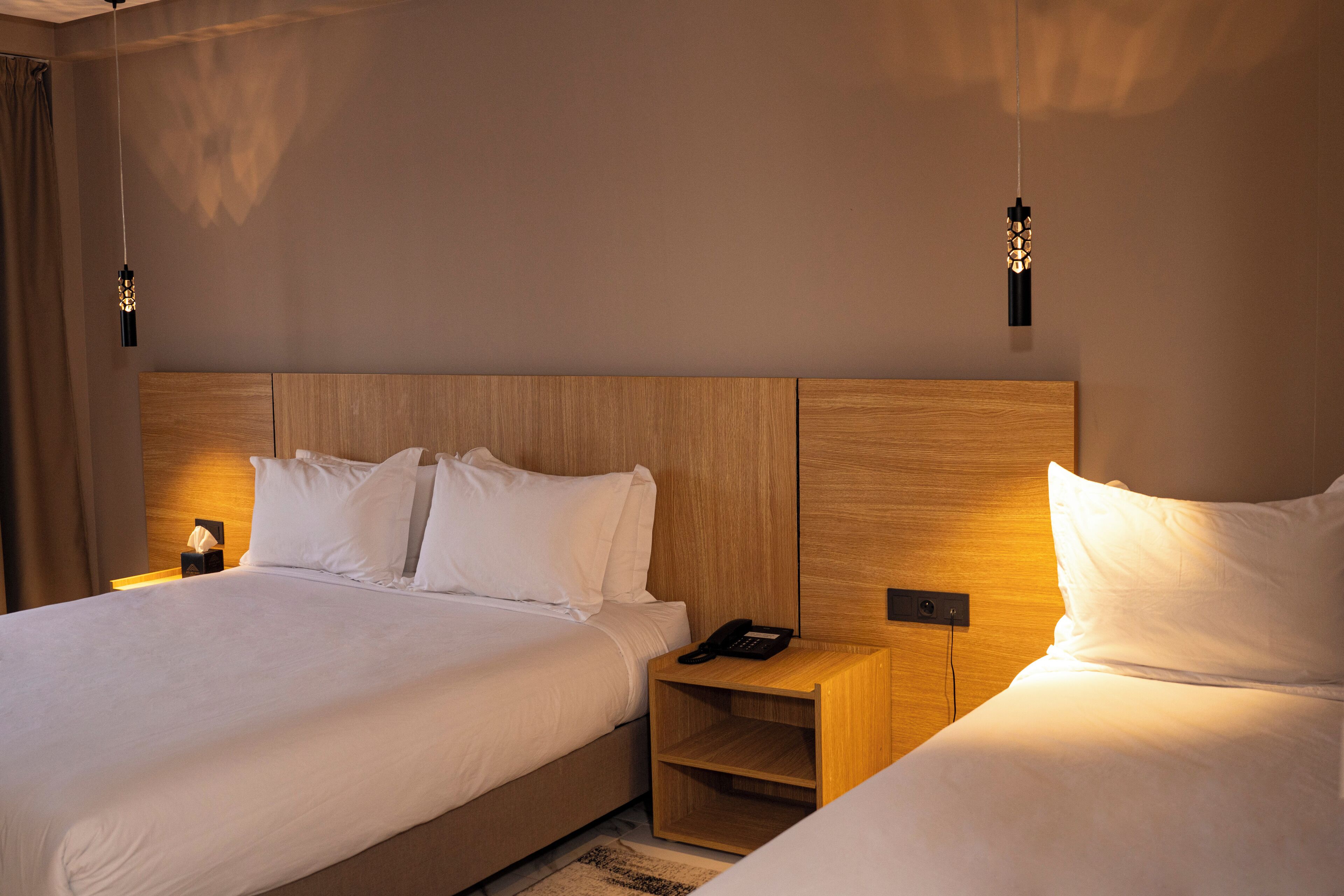 Quarto de hotel com uma cama grande, uma mesinha de cabeceira com um telefone e uma mesa lateral com abajur.