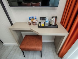 Quarto de hotel com secretária de madeira, um banco estofado marrom, televisão de tela plana e minibar com lanches e chaleira.