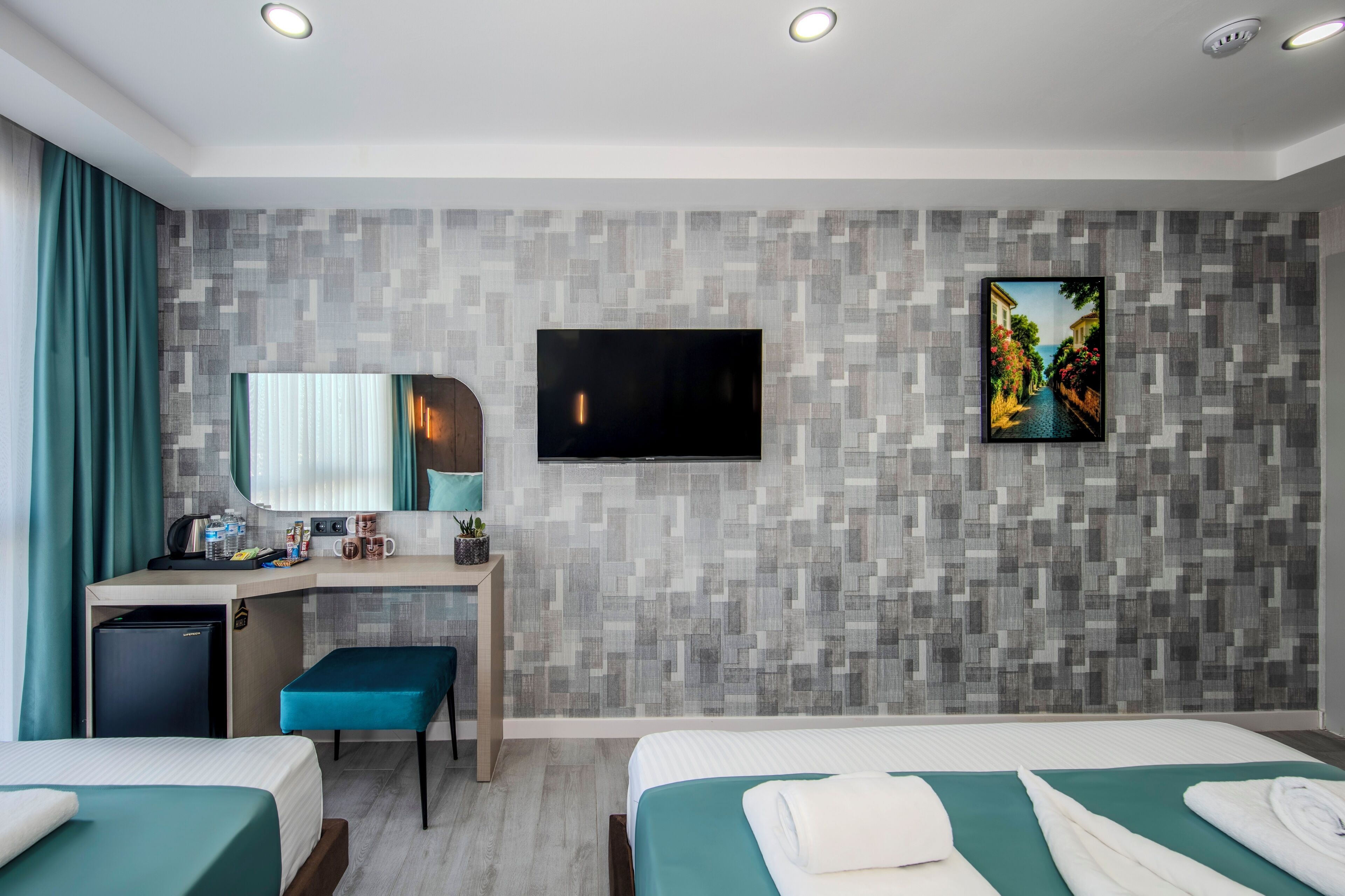 Quarto de hotel com duas camas, uma escrivaninha com computador, uma televisão e um painel de parede revestido de azulejos.