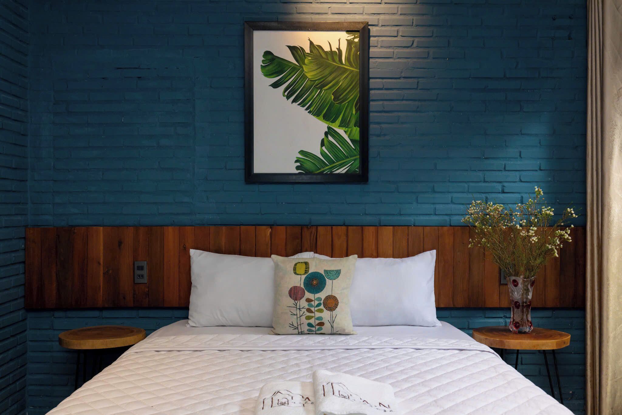 Quarto de hotel com cama, cabeceira de madeira, parede de tijolos azuis, quadro com folhagem tropical, almofada decorativa, mesa redonda pequena com vaso de flores e mesa de cabeceira com luminária.