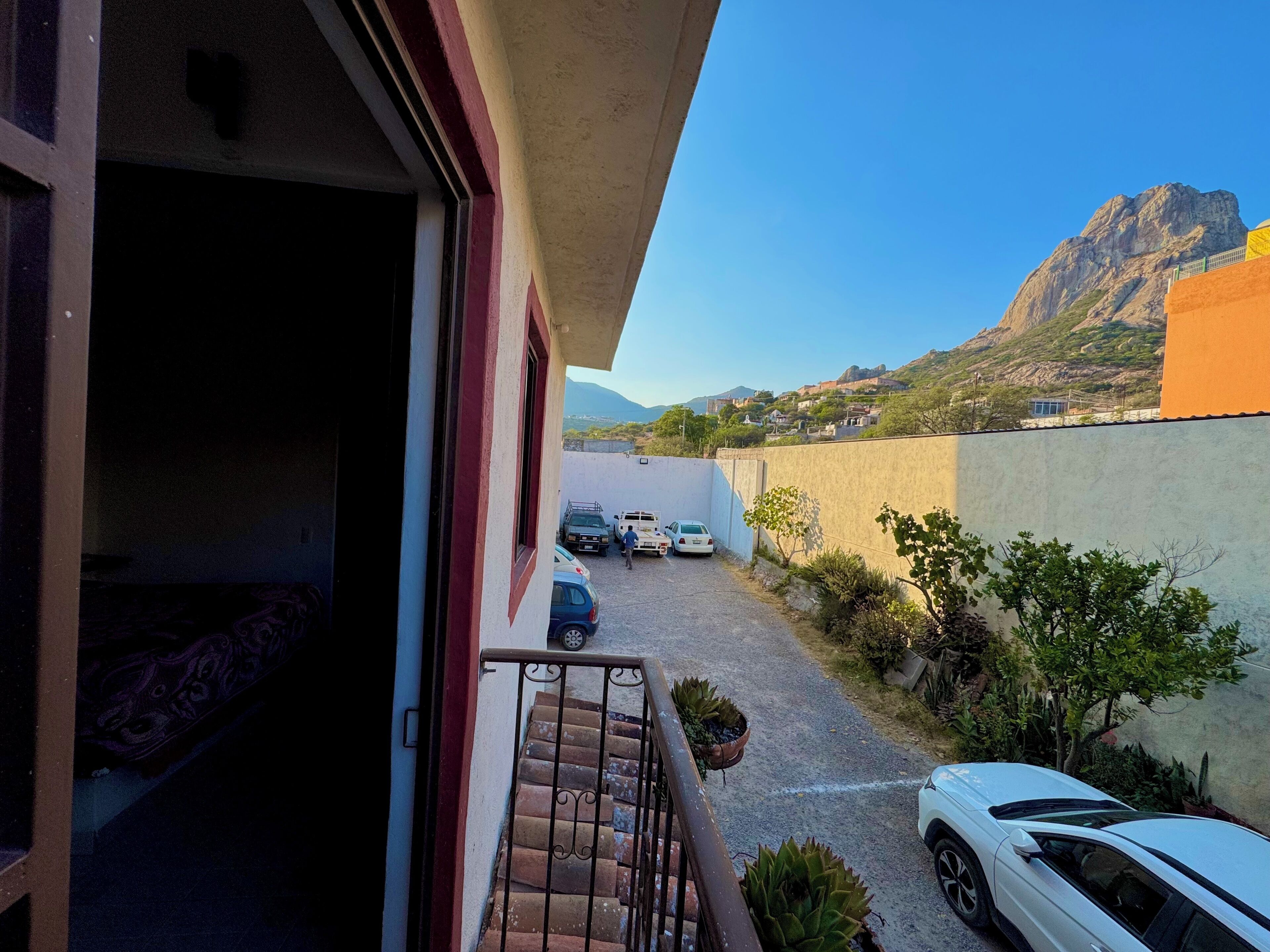 Uma varanda com vista para um parque de estacionamento, uma montanha ao fundo e um edifício com fachada vermelha e bege.