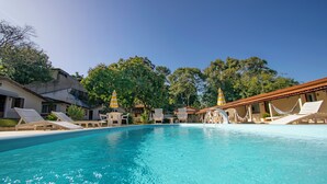 Piscina azul cristalina com espreguiçadeiras e guarda-sóis, rodeada por árvores e edifícios.