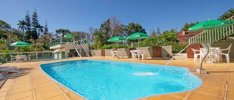 Piscina com deck ladrilhado, rodeada por móveis de exterior e guarda-sóis verdes.