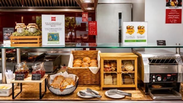 Buffet de café da manhã com diversos alimentos, uma máquina de café e uma placa indicando o cardápio.