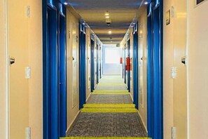 Corredor de hotel com portas azuis, chão coberto por carpete e um extintor de incêndio.