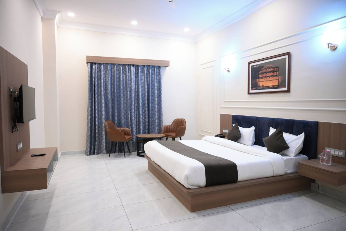 Quarto de hotel com uma cama grande, uma escrivaninha, duas cadeiras, uma televisão e uma janela com cortinas.