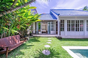 Uma casa com telhado azul, alpendre coberto, piscina e um gramado bem cuidado com pedras no caminho.