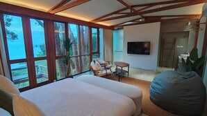 Quarto com uma cama grande, área de estar com uma cadeira e mesa, e vista para o exterior através de janelas amplas.