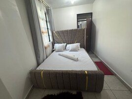 Uma cama bem arrumada com lençóis e almofadas brancas, uma cabeceira cinzenta e um tapete vermelho que leva a uma porta.