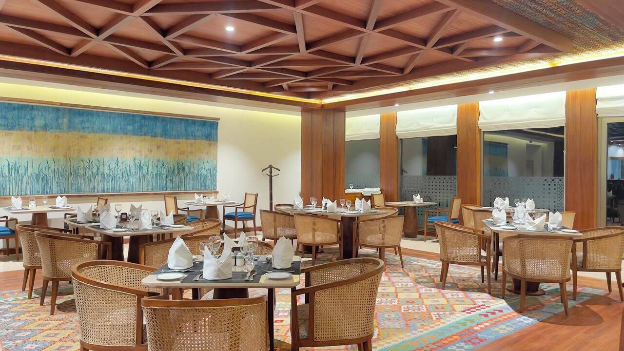 Um restaurante com várias mesas e cadeiras, teto com padrão geométrico e uma grande pintura abstrata na parede.