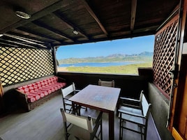 Quarto com teto de madeira, uma mesa com cadeiras, um sofá estampado e vista para um lago e montanhas.