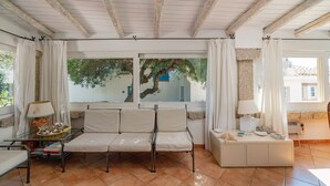 Um quarto aconchegante com sofá, uma pequena mesa e um armário. Há janelas com cortinas, uma árvore visível lá fora e piso de azulejos.