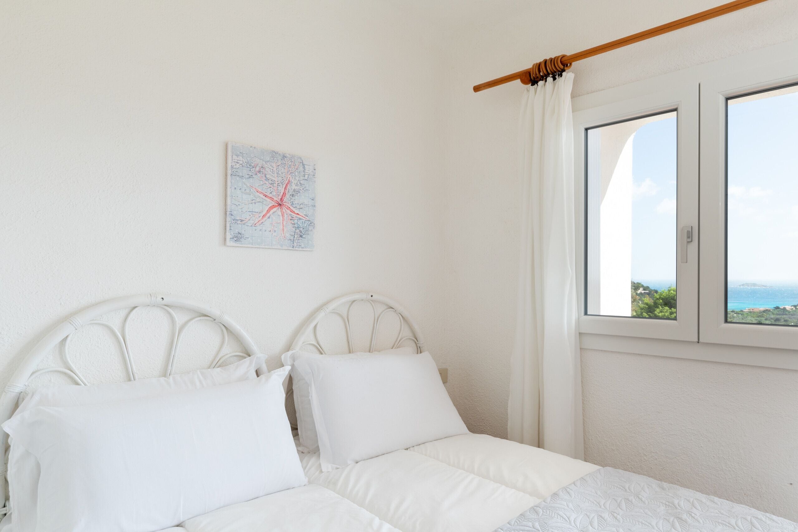 Quarto com cama, cortinas brancas, janela com vista e uma obra de arte na parede.