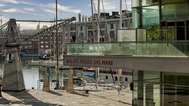 Um estaleiro com barcos atracados, uma ponte ao fundo e um edifício com a placa 