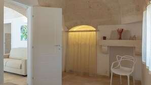 Quarto com porta branca, cadeira branca, área de chuveiro com cortina e um pequeno armário com frascos.