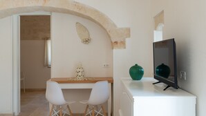 Quarto com uma secretária branca, duas cadeiras brancas, uma televisão sobre um armário branco e um objeto decorativo na parede.