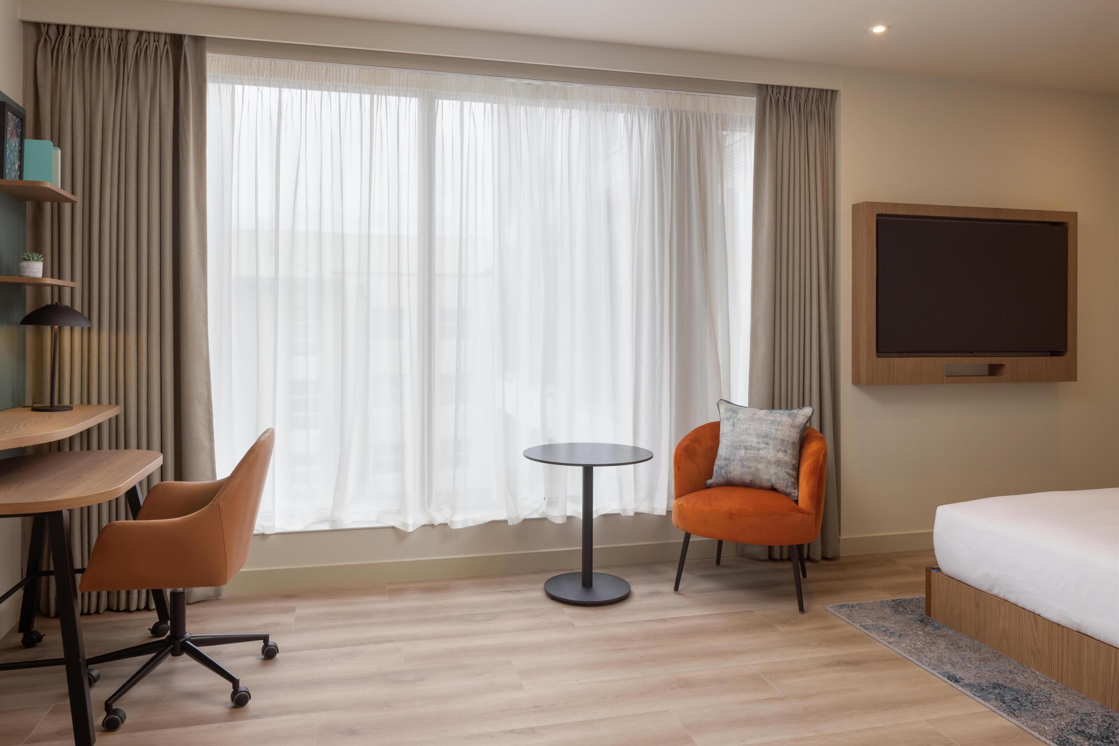 Quarto de hotel com uma cama, uma escrivaninha com cadeira, uma mesa redonda pequena e uma televisão fixada na parede.