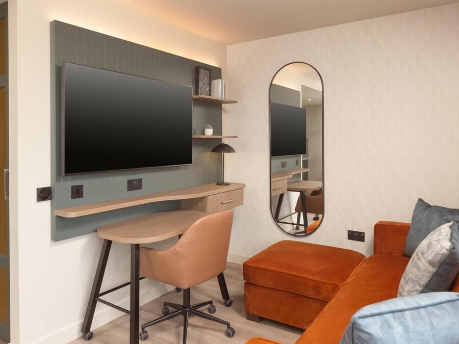 Quarto moderno com televisão na parede, escrivaninha com cadeira, sofá com almofadas e um espelho grande.