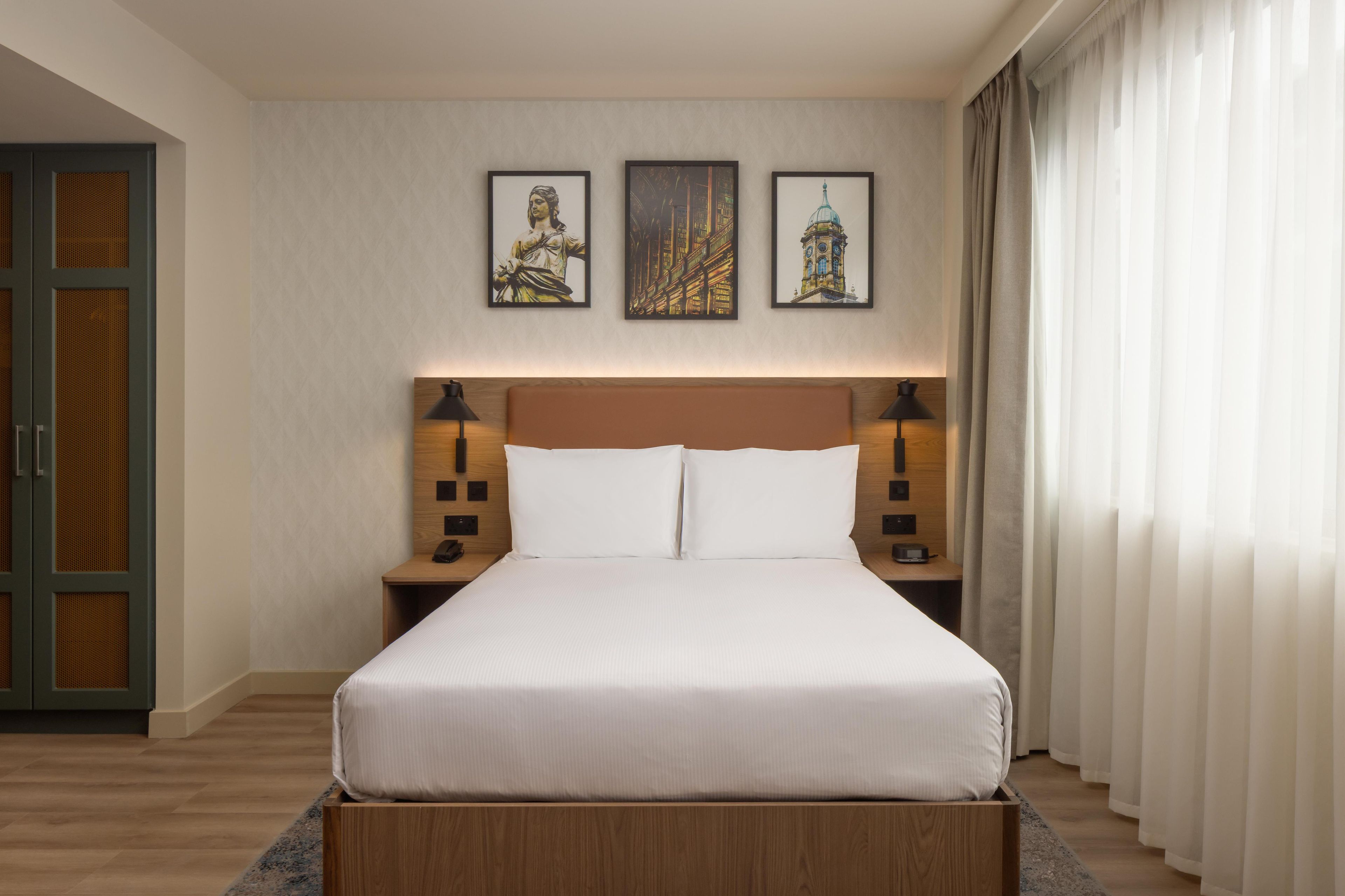 Uma cama bem arrumada com cabeceira de madeira, duas mesas de cabeceira com luminárias e três quadros na parede.
