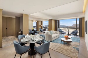 Quarto de hotel moderno com área de jantar, sala de estar com sofá e poltronas, e varanda com vista para a cidade.