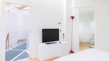 Quarto moderno com cama, TV sobre cômoda branca, abajur vermelho e vista para varanda e banheiro.