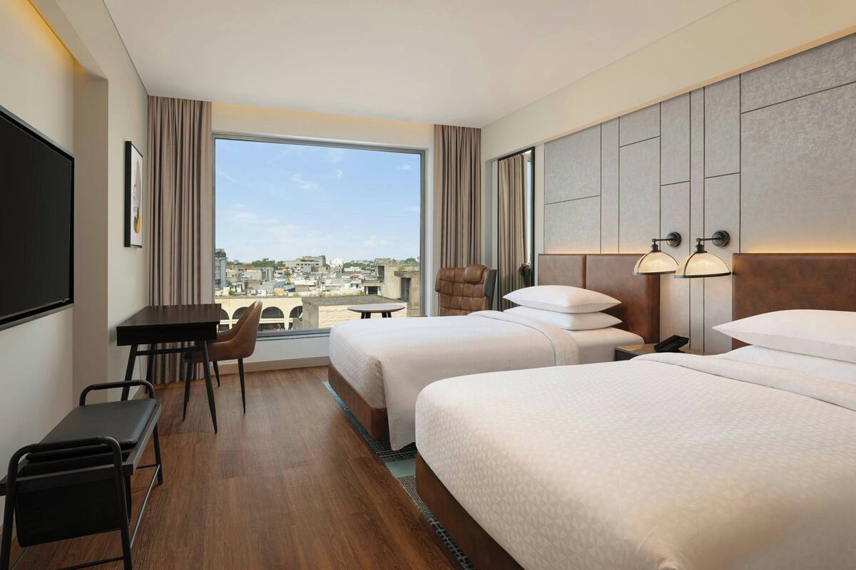 Quarto de hotel com duas camas, uma escrivaninha, uma cadeira e uma janela grande com vista para a cidade.
