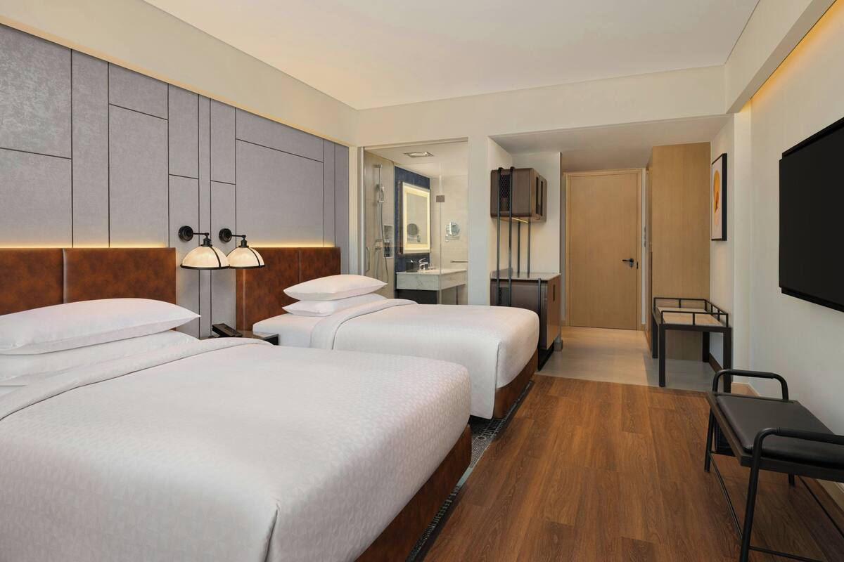 Quarto de hotel com duas camas, banheiro, televisão e escrivaninha.