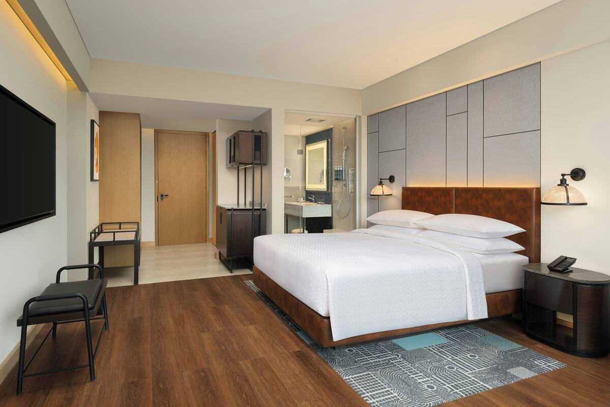 Quarto de hotel moderno com uma cama grande, mesas de cabeceira, escrivaninha e banheiro visível através de uma porta aberta.