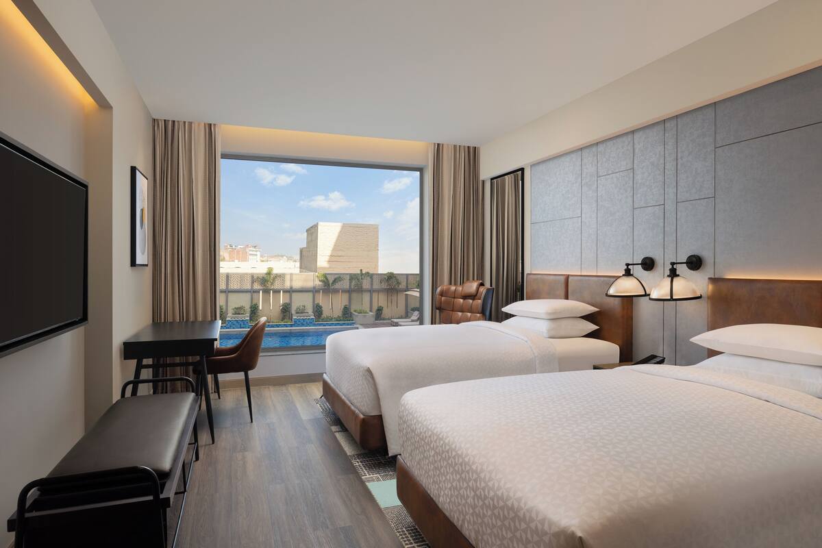 Quarto de hotel com duas camas, uma escrivaninha, uma cadeira e uma televisão. Há uma janela com vista para edifícios e uma piscina.