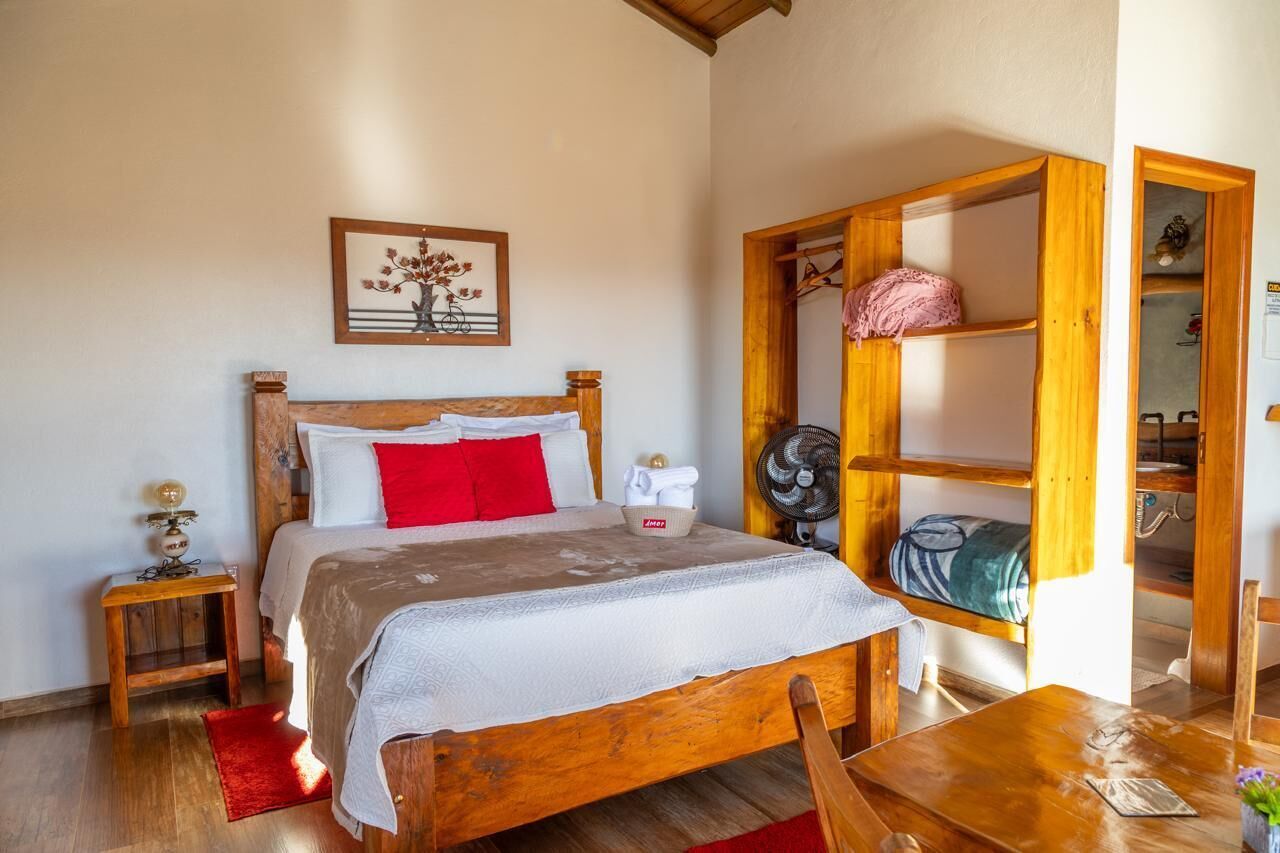 Uma cama de madeira com colcha branca e vermelha, uma pequena mesa de cabeceira com abajur e uma cadeira de madeira ao lado de uma mesa de madeira.