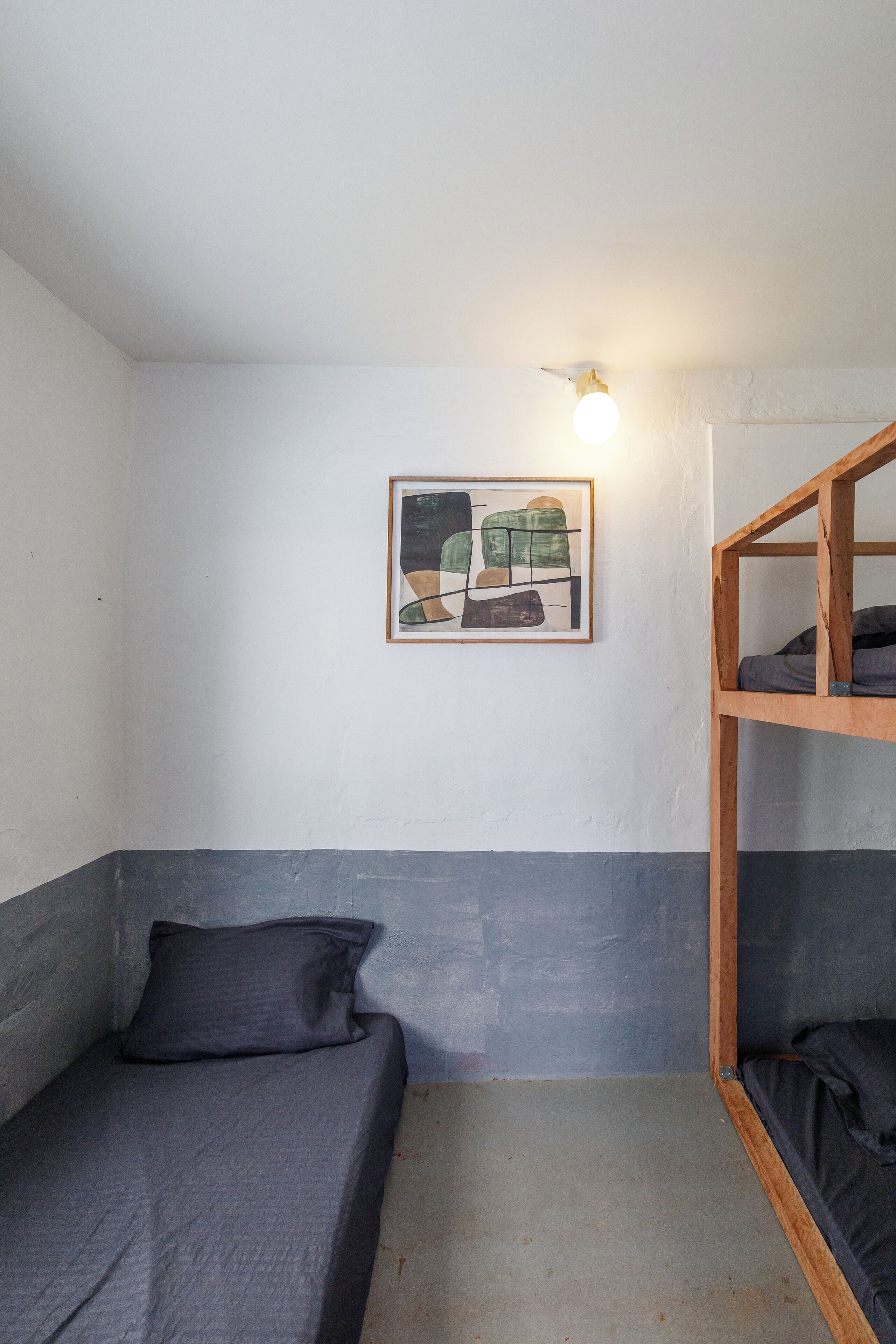 Quarto pequeno com uma cama de solteiro e um beliche no lado direito, uma pintura abstrata emoldurada na parede e um aparelho de luz sobre a cama.