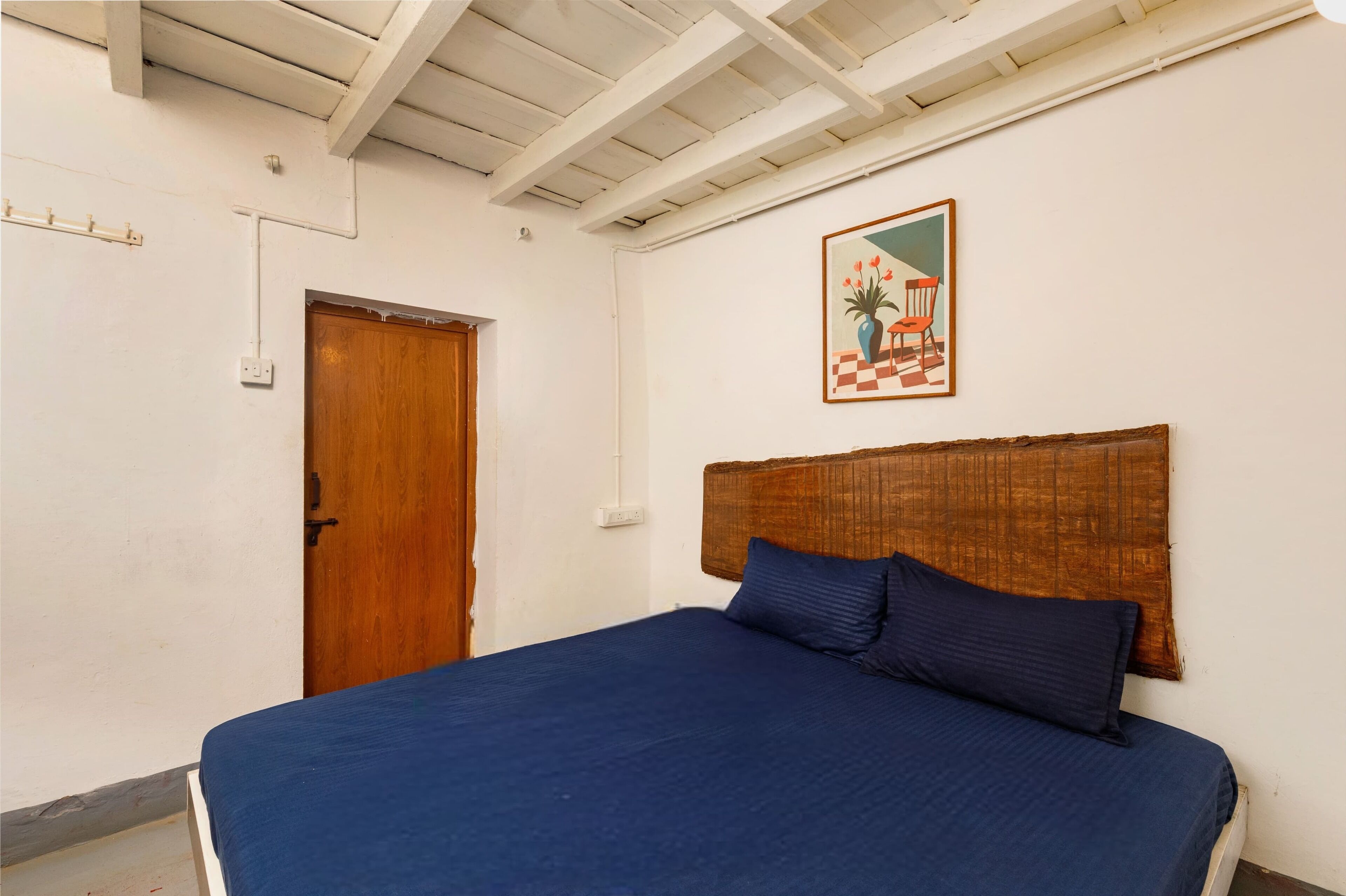 Quarto com porta de madeira, cama com colcha azul, cabeceira com textura trançada e pintura na parede de uma cadeira e planta.