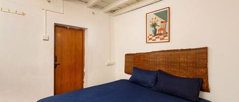 Quarto com porta de madeira, cama com colcha azul, cabeceira com textura trançada e pintura na parede de uma cadeira e planta.