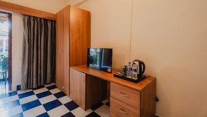 Quarto compacto com uma escrivaninha de madeira, televisão, chaleira e bandeja com garrafas de água.