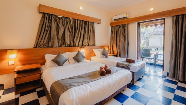 Quarto de hotel com piso xadrez, uma cama grande com duas almofadas, mesa de cabeceira e janela com cortinas.