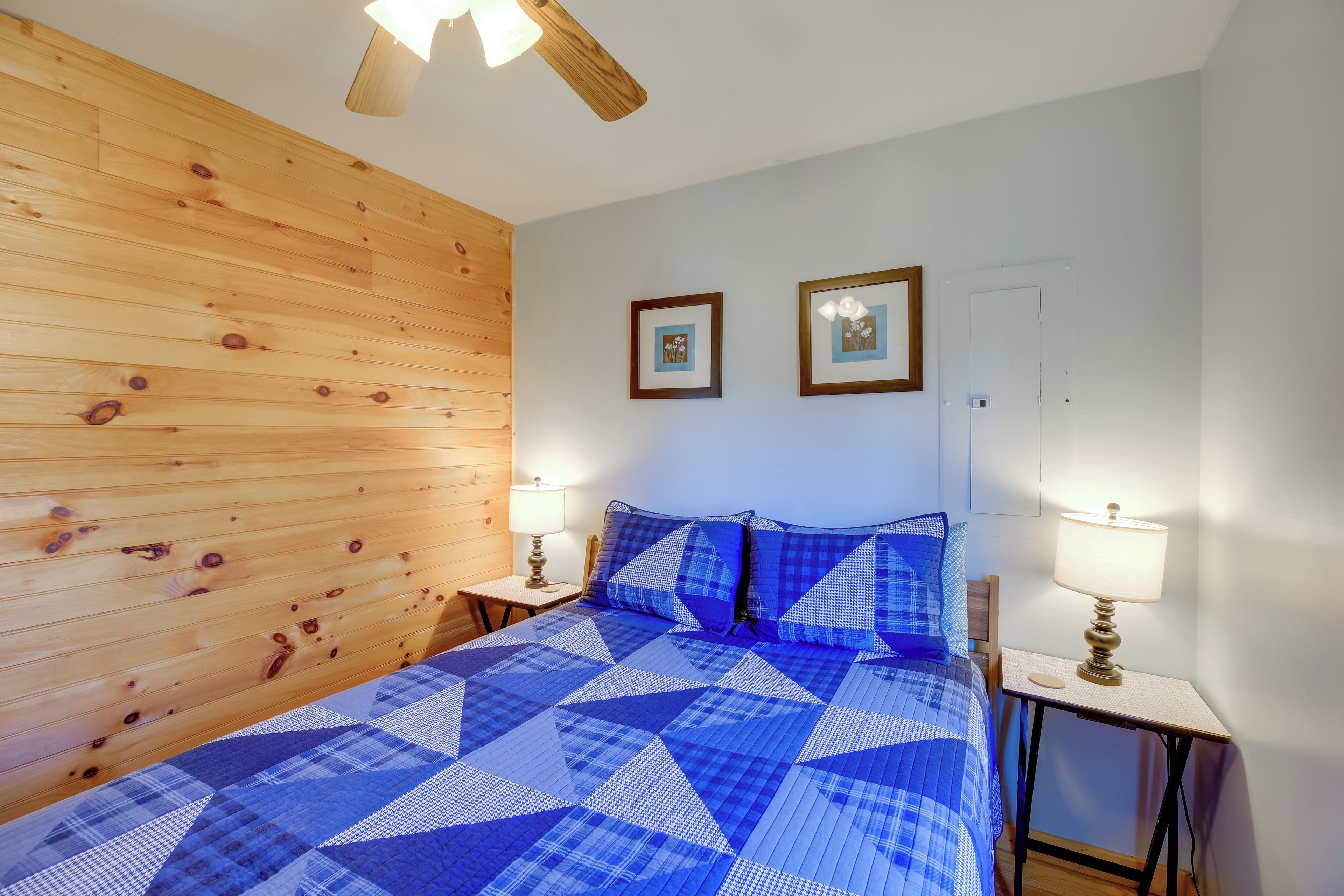 Quarto com parede de madeira, cama com colcha xadrez azul, duas imagens emolduradas na parede e duas luminárias de cabeceira.