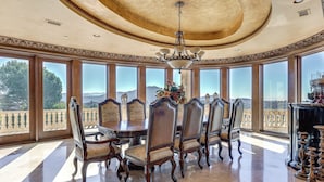 Uma sala de jantar com uma mesa grande e cadeiras, janelas amplas com vista e um candelabro.