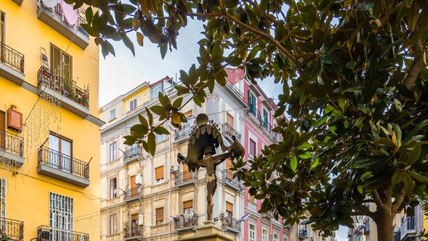 Praça com plantas em vasos, uma estátua e um edifício com a placa 