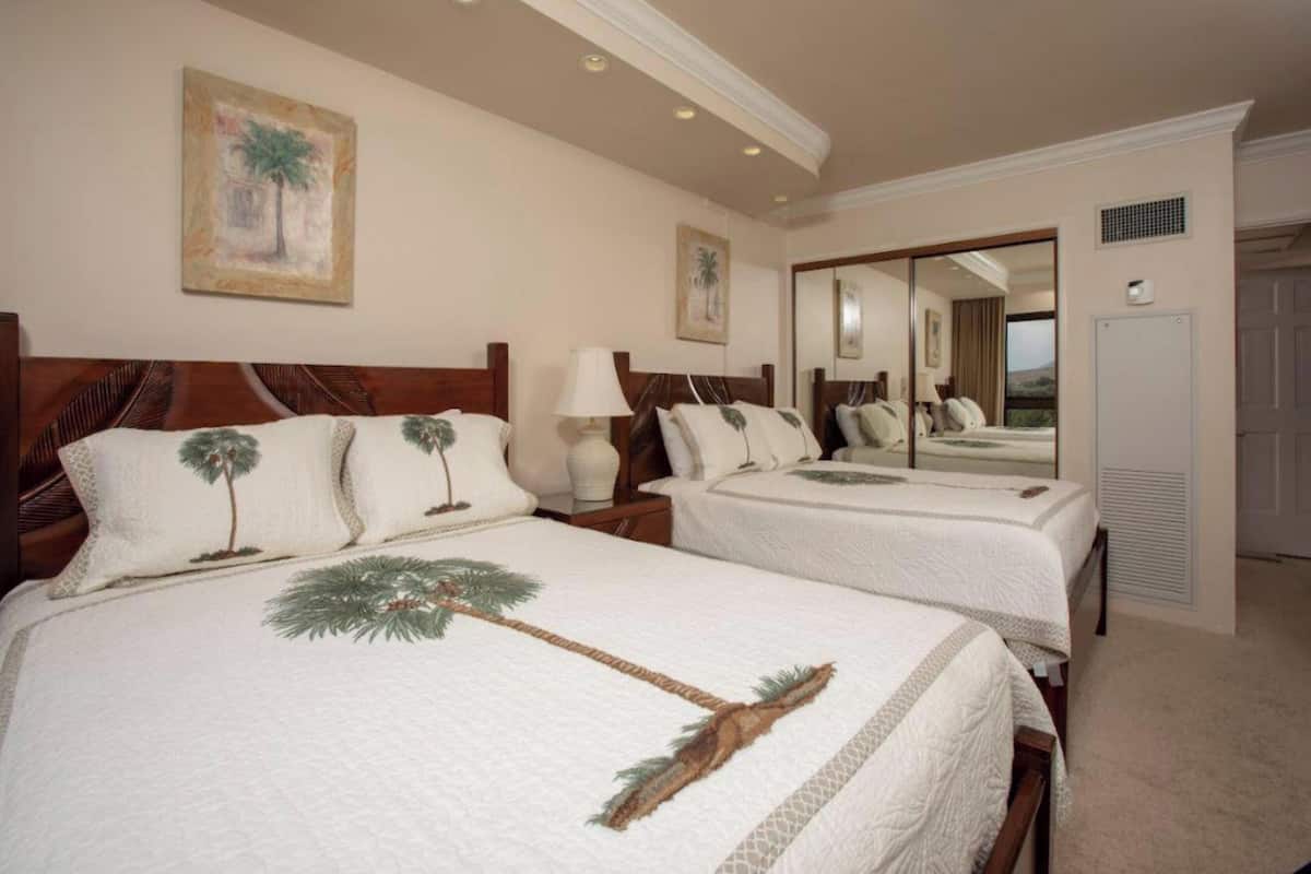 Quarto de hotel com duas camas, cada uma com desenho de palmeira, uma mesa de cabeceira com abajur, um espelho e uma porta com respiro.