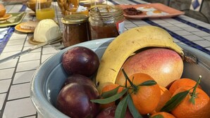 Uma mesa posta com frutas, doces e bebidas, com uma parede de pedra e uma chaminé ao fundo.