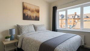 Quarto com cama, mesa de cabeceira com abajur, uma pequena planta e janela com vista para edifícios.