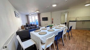 Uma sala de jantar com uma mesa posta para quatro pessoas, rodeada por cadeiras brancas, um sofá e uma televisão fixada na parede.