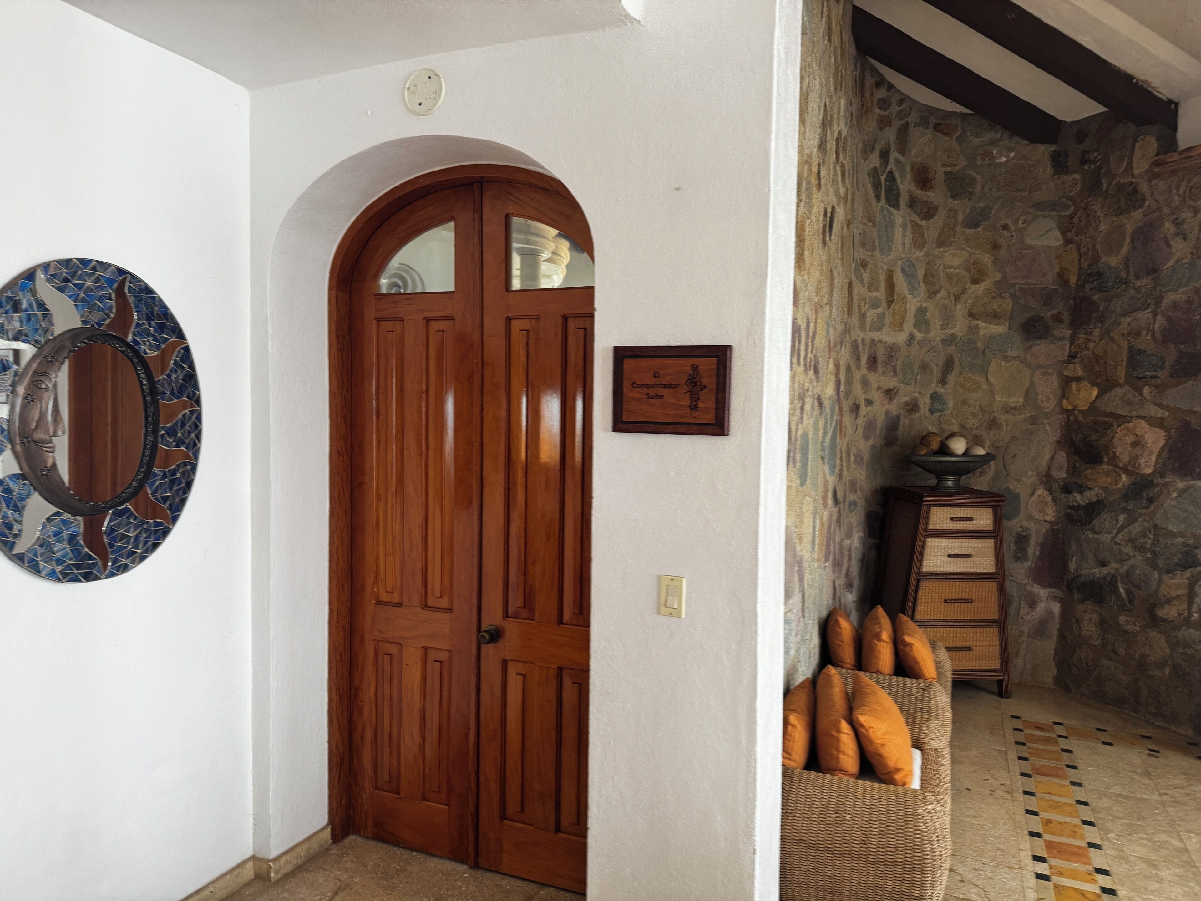 Uma porta de madeira com um painel de vidro, um espelho fixado na parede, uma placa emoldurada, uma parede de pedra, um sofá de vime com almofadas e um chão de azulejos.