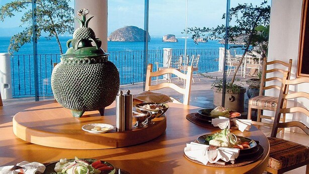 Mesa de jantar com pratos de comida, um vaso decorativo e cadeiras em um quarto com vista para o mar.