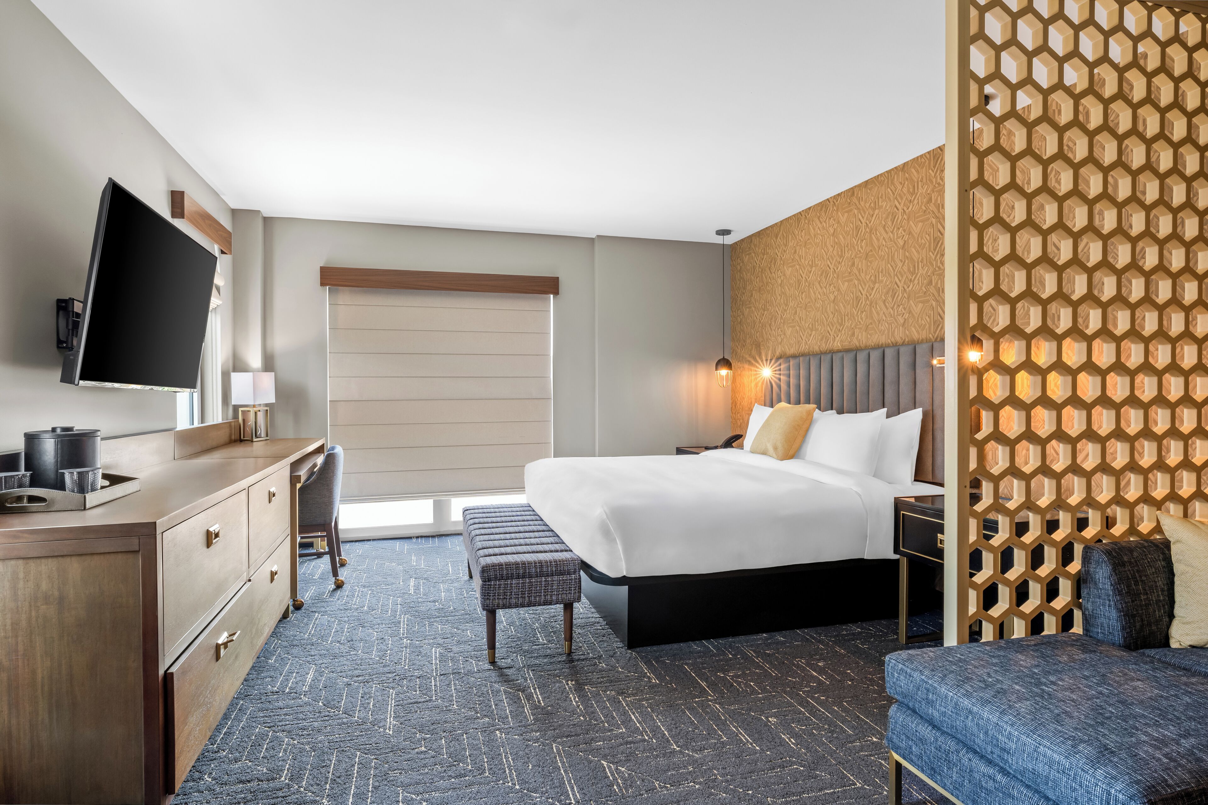 Quarto de hotel com cama, mesa de cabeceira, televisão, escrivaninha, cadeira e parede com padrão.