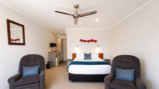 Quarto de hotel com cama, duas poltronas, ventoinha no teto, espelho e uma pequena cozinha equipada.