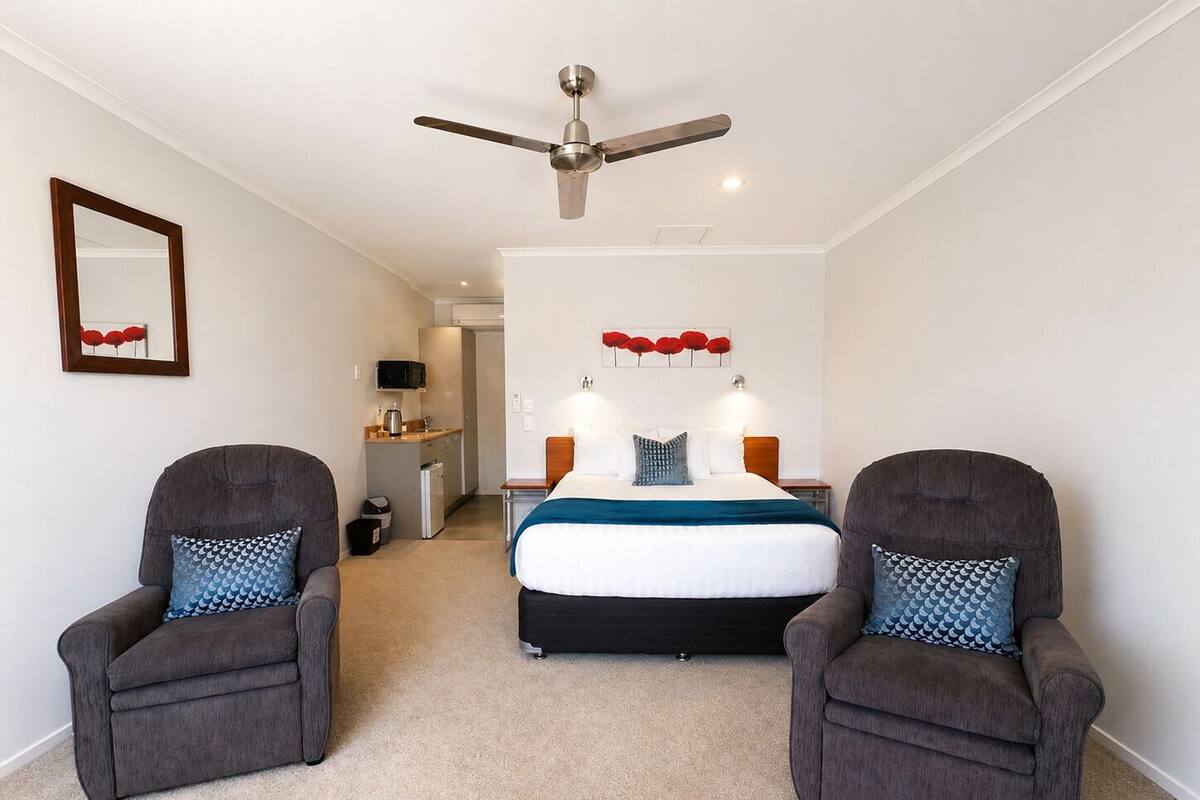 Quarto de hotel com cama, duas poltronas, ventoinha no teto, espelho e uma pequena cozinha equipada.